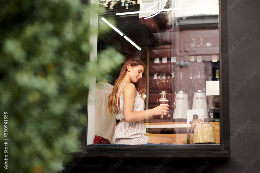 Young blond woman, wearing white overall, sitting by huge glass window ...