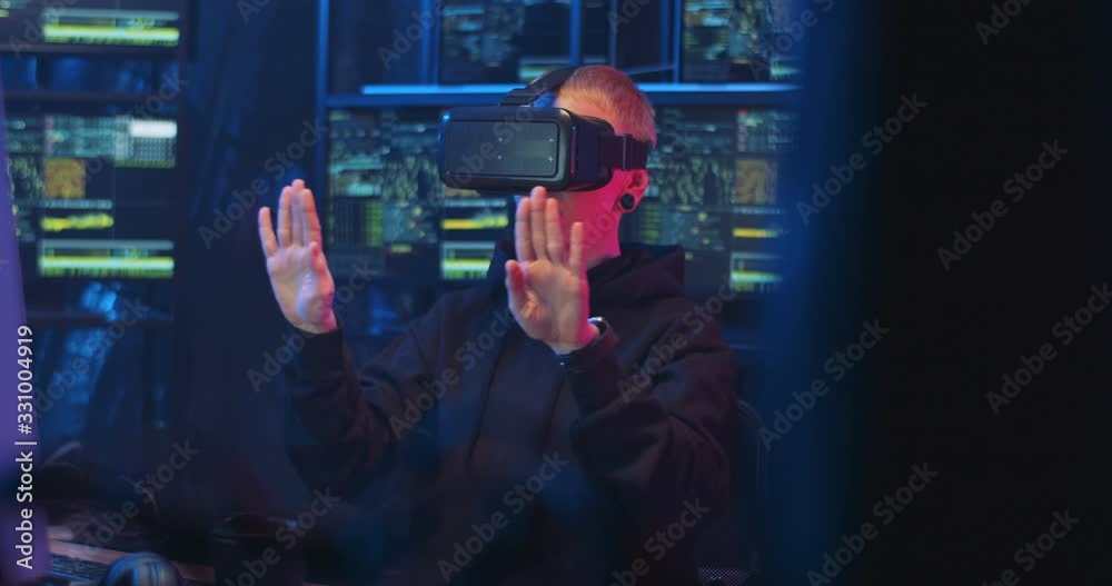 Caucasian female hacker in VR glasses sitting in darkness in monitoring room full of computers ...