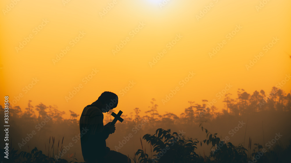 young human kneeling down and praying with christian cross at sunset ...