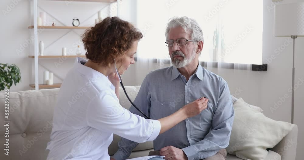 Female doctor listening old adult patient with stethoscope during home ...