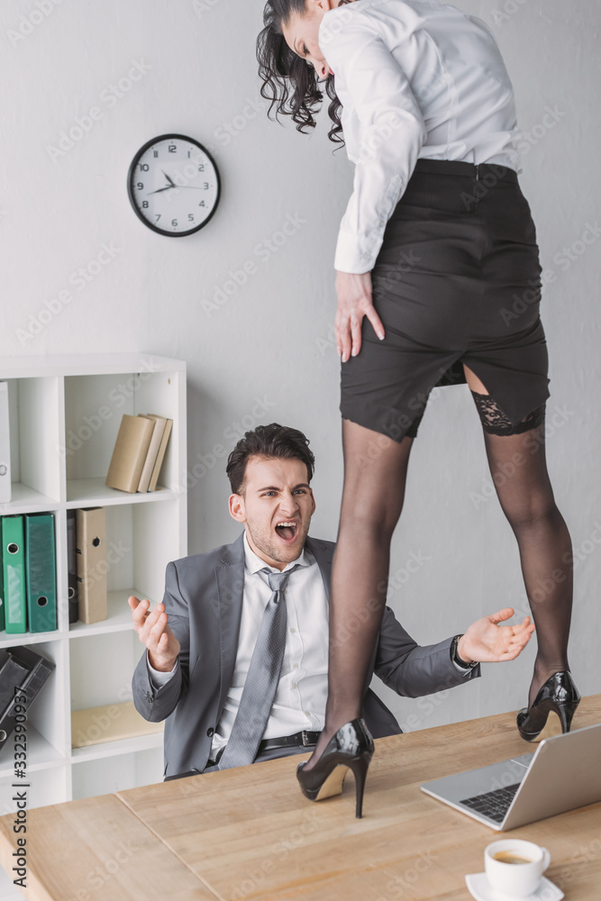 angry businessman screaming while looking at sexy secretary standing on desk in high heeled ...