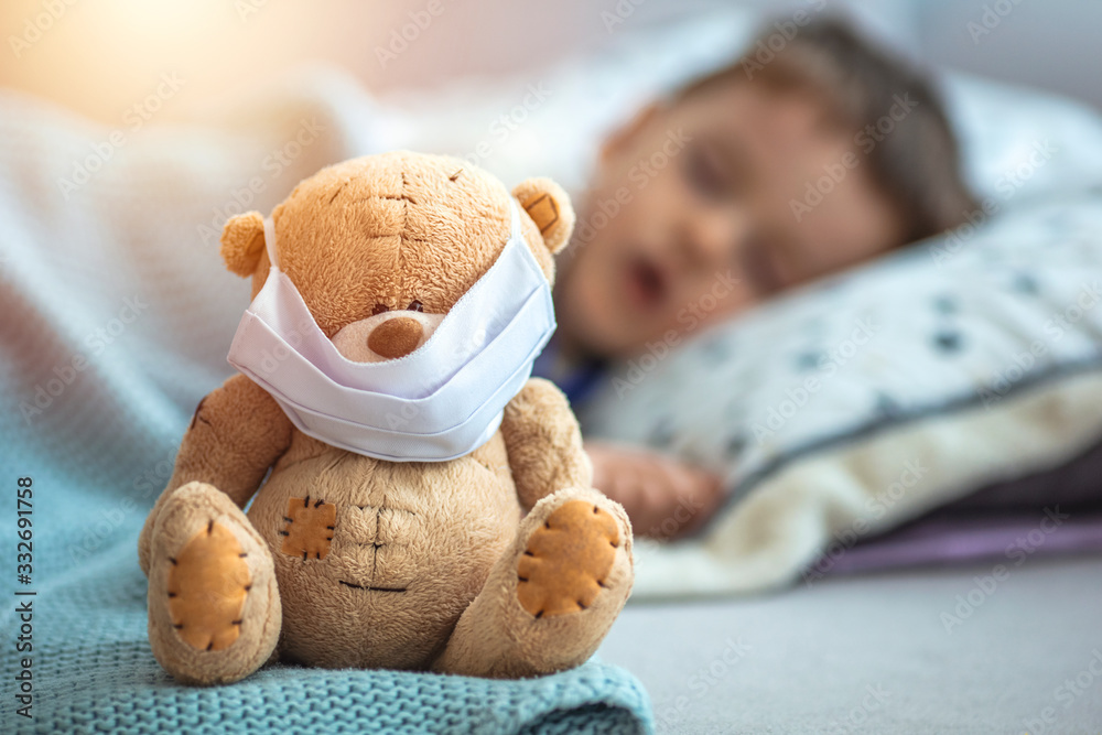 Child in home quarantine at the bed, sleeping, with medical mask on his sick teddy bear, for ...