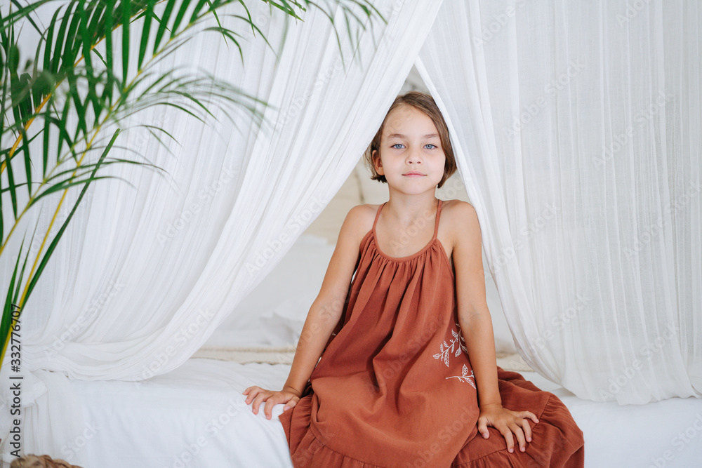 Portrait of a pretty little girl in a dress sitting on a double bed ...