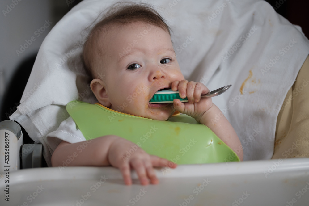 Feeding. Messy smiling baby eating with a spoon in high chair. Baby's ...
