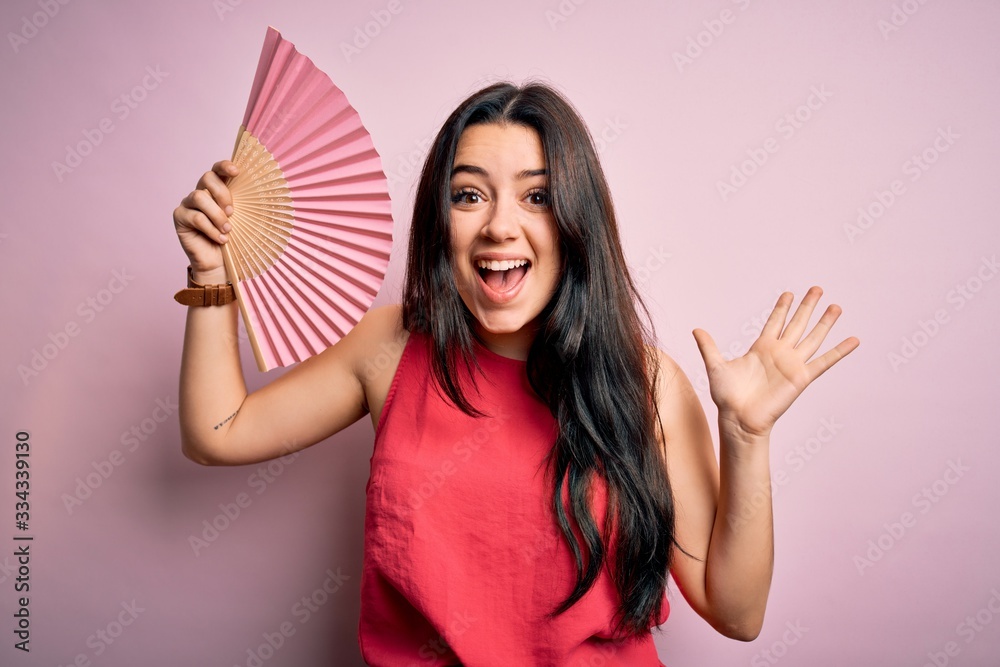 Young brunette woman holding hand fan for fresh air over pink isolated ...
