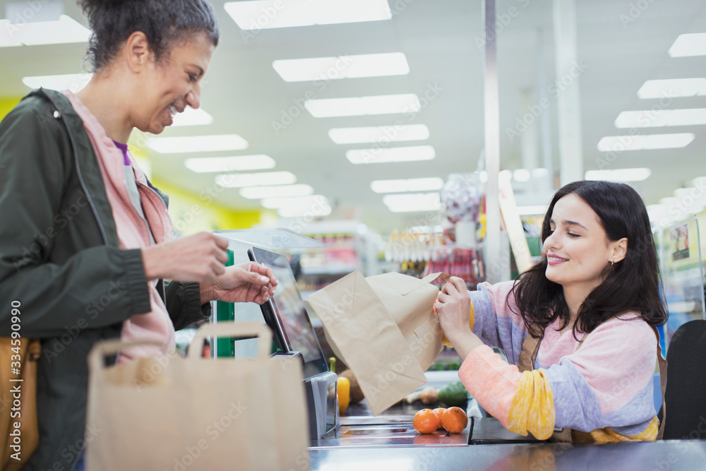 Female cashier helping customer bag groceries at supermarket checkout ...