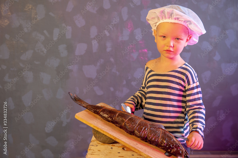 Little blond boy in cook's hat shows big smoked fish. Child tries fish ...