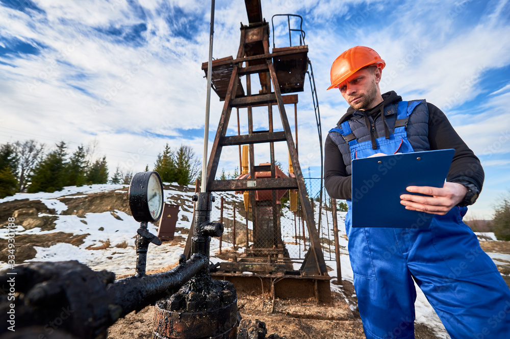 Serious oil man in work overalls and helmet holding clipboard and ...