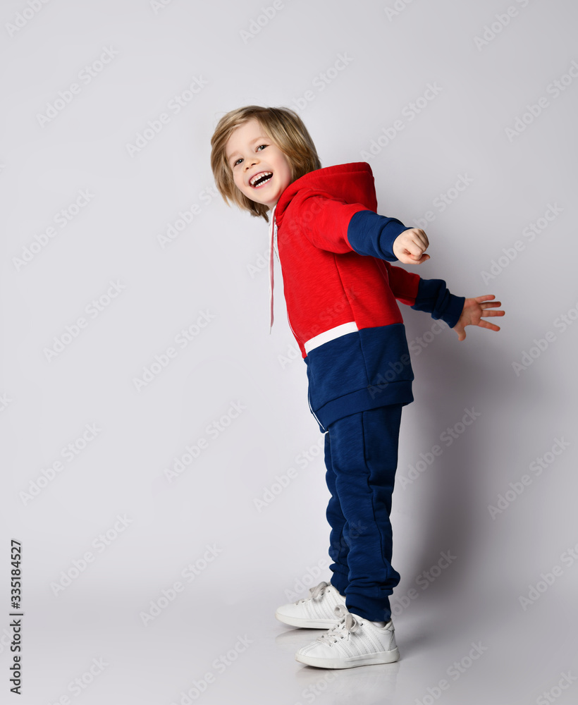 Full growth portrait of frolic blond kid boy in red and blue sport suit standing sideways to us ...