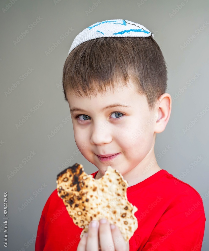 Cute Caucasian child in a white kippah cap eating shmura matzo, a piece ...