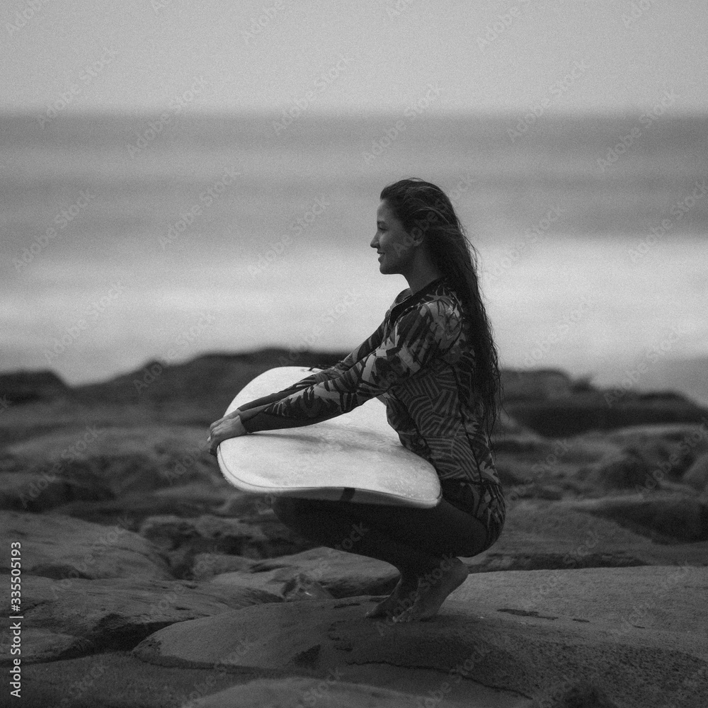 Monochrome. Surfer girl sitting on the rock. Blue longboard. Surfing lifestyle. Young woman with ...