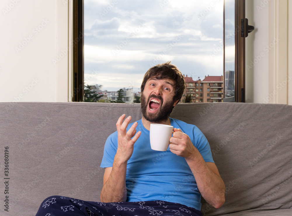 young cool man sitting on a sofa at living room with a cup. Angry ...