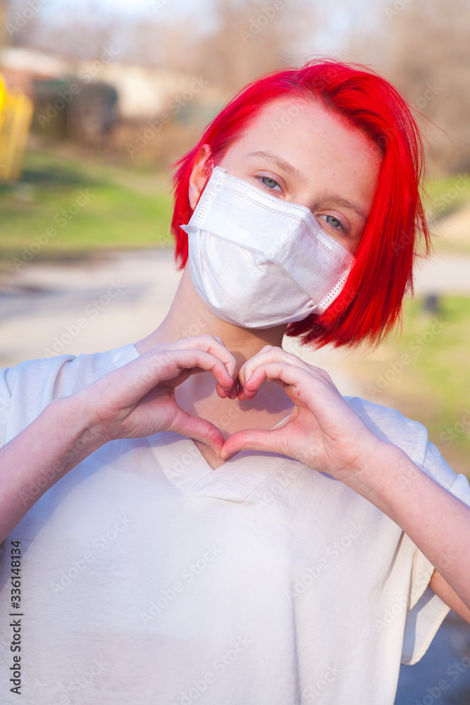Red hair teenage girl wearing a medical mask shows the heart symbol ...