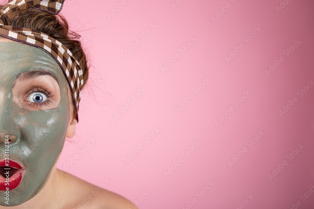 Surprised girl in a clay mask on her face standing on a pink background ...