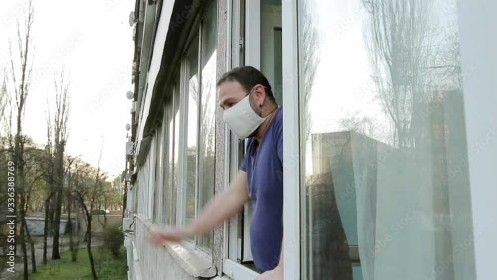 A man in a medical mask opens a window waving hello, spreads his hands ...