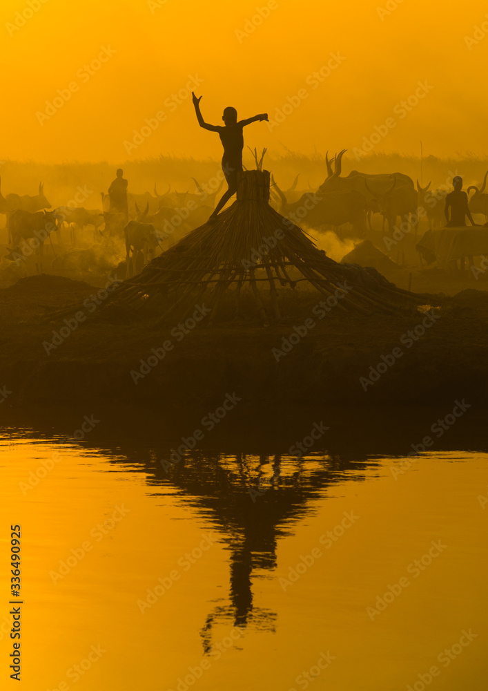 A Mundari tribe boy on a hut roof mimics the position of horns of his ...