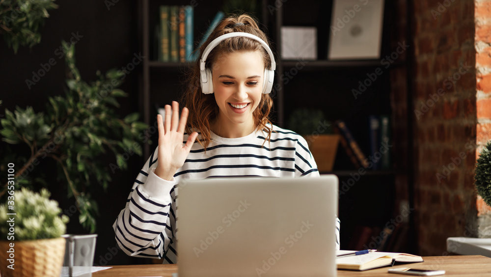 Cheerful woman in headphones greeting friend while talking on laptop at ...