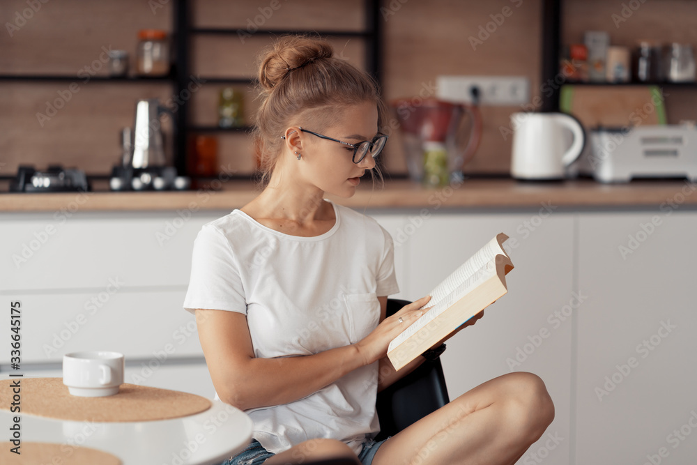 Cute girl is drinking coffee and reading a book at the kitchen table at ...
