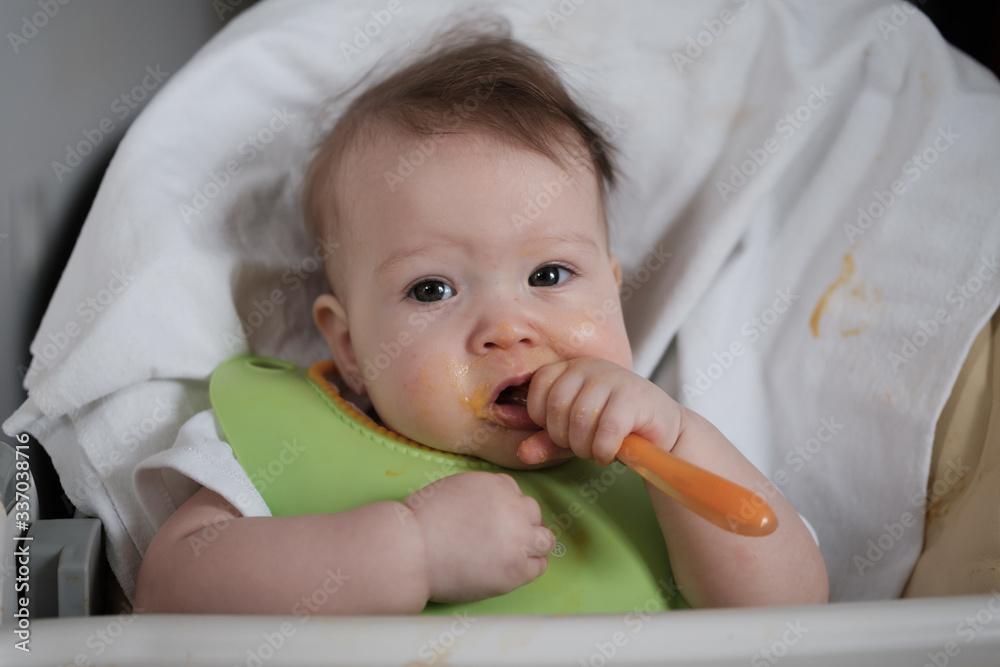 Feeding. Messy smiling baby eating with a spoon in high chair. Baby's ...