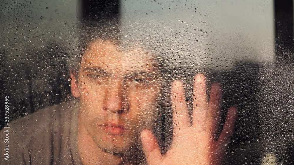 Sad Guy Caucasian appearance looks through a wet window after a rain. Close-up. On him falls the ...