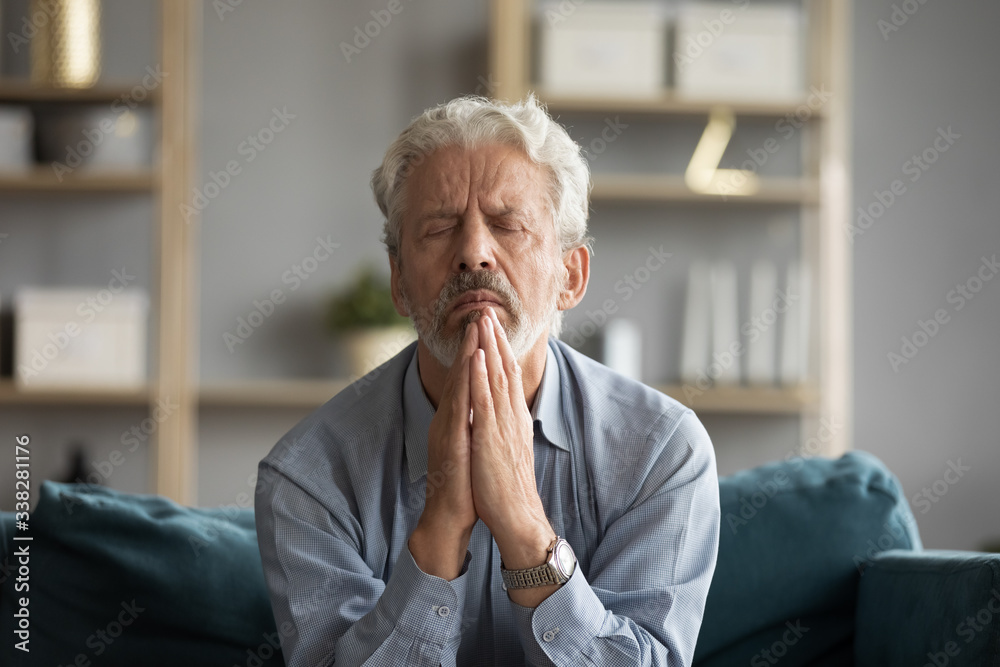 Sad older man with closed eyes praying with hope, sitting on couch at ...
