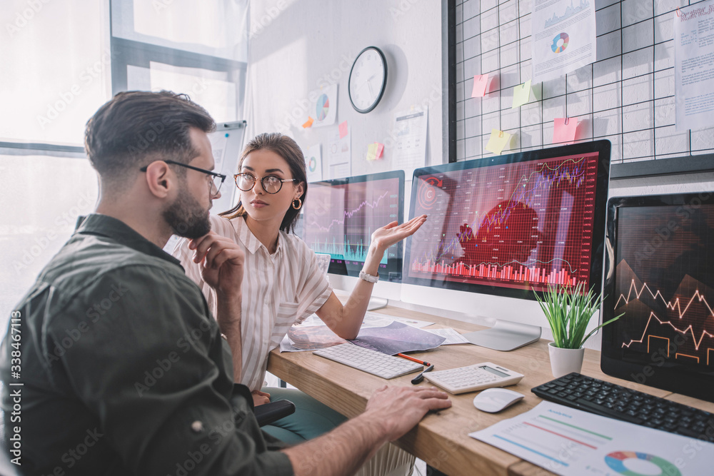 Data analyst pointing with hand at charts on computer monitor to colleague at table in office ...