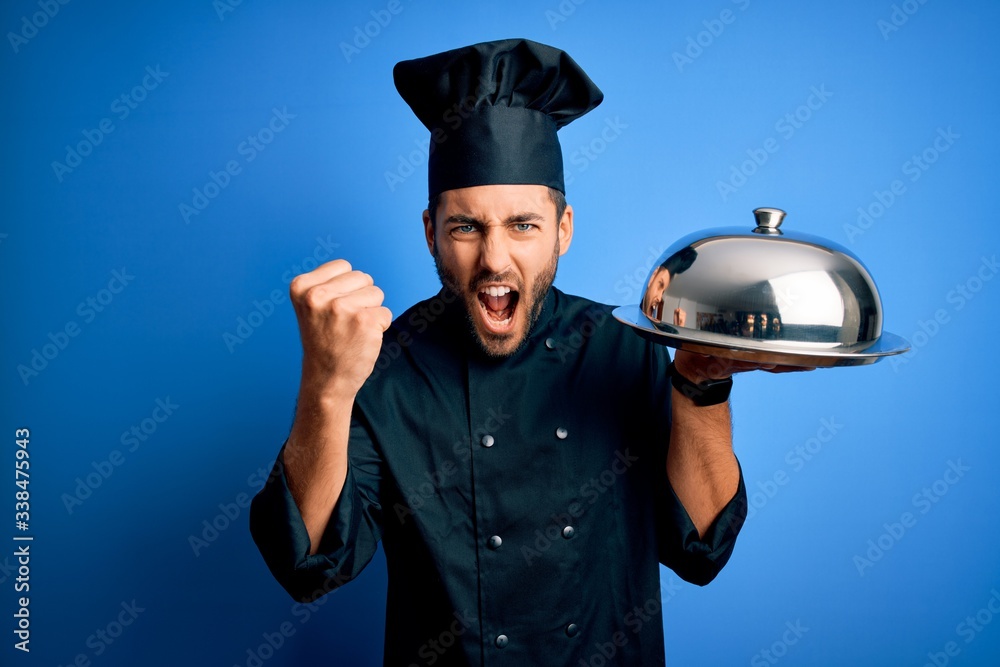 Young cooker man with beard wearing uniform holding tray with dome over ...