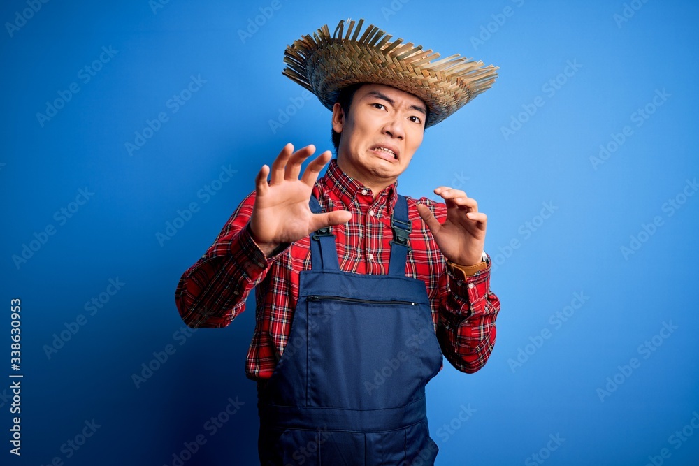 Young handsome chinese farmer man wearing apron and straw hat over blue ...