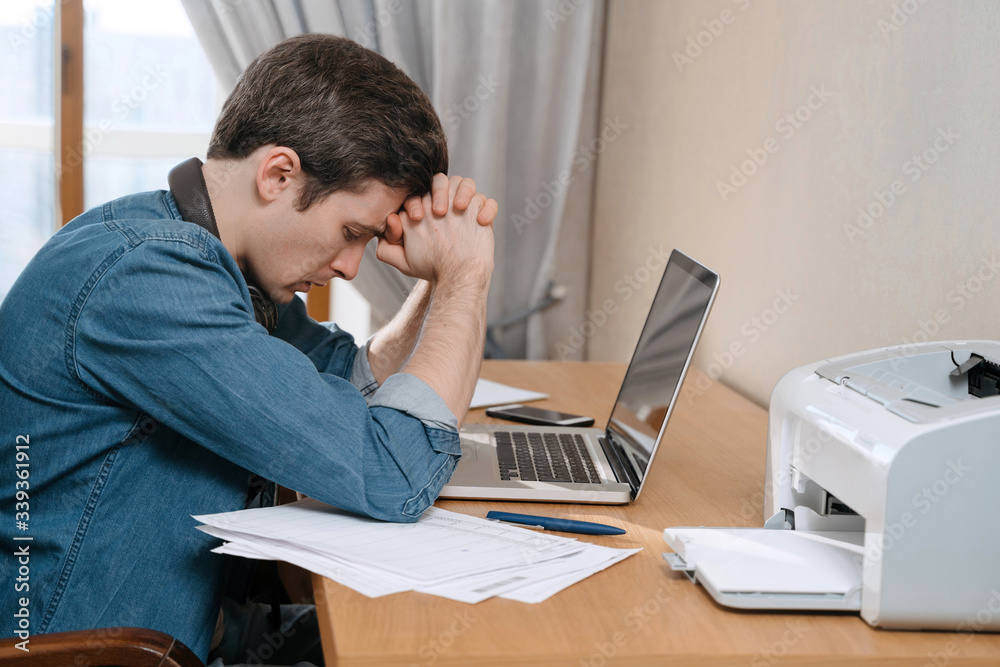 Confused tired young man sitting at his desk with laptop, hands folded ...