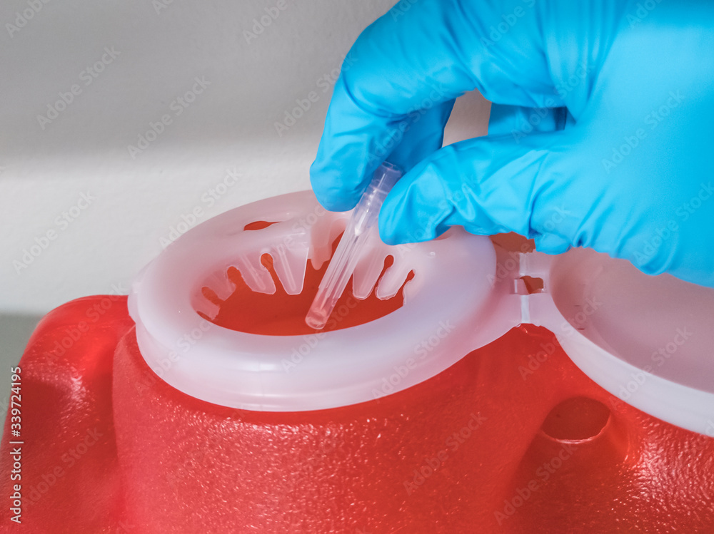gloved hand dropping a used needle into a bright red sharps container ...