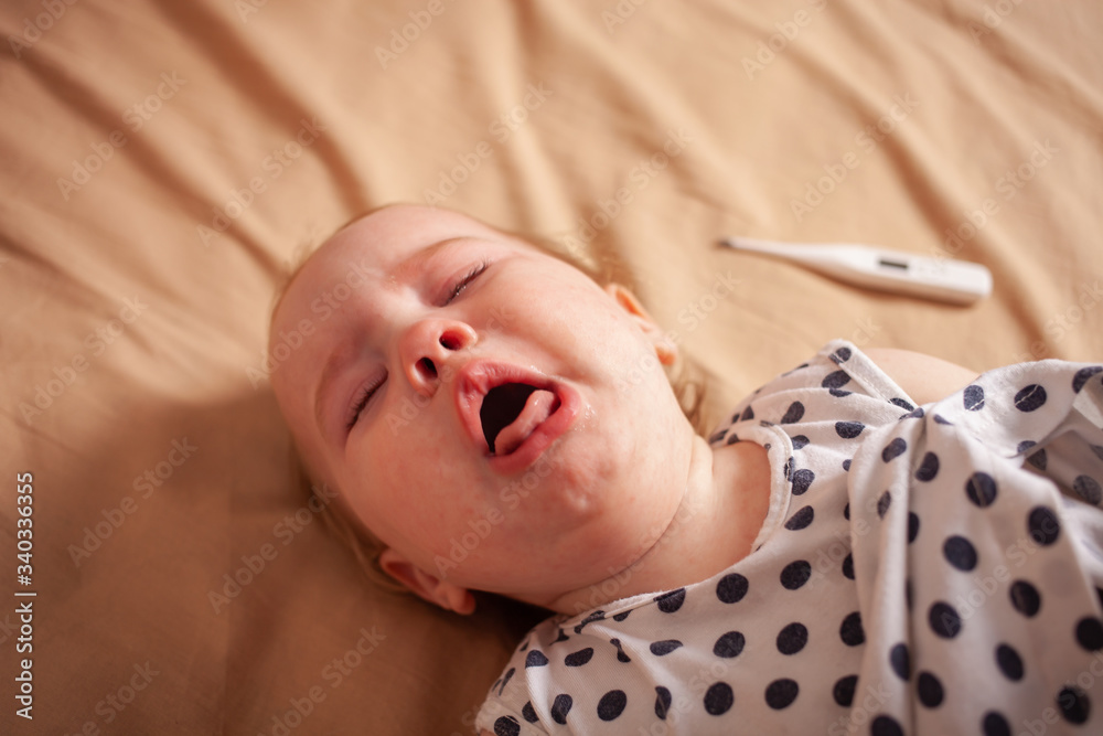 A sick child lies on the bed and screams. Thermometer next to a beige ...