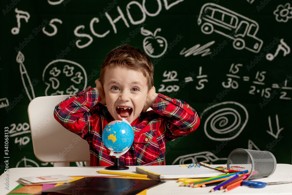 Emotional school boy sitting on the desk with many school supplies ...