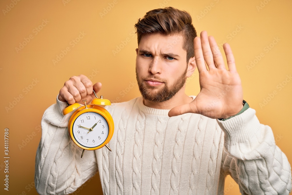 Young blond man with beard and blue eyes holding alarm clock over ...