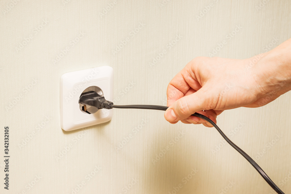 A person’s hand pulls an electrical cord plugged into a power outlet ...