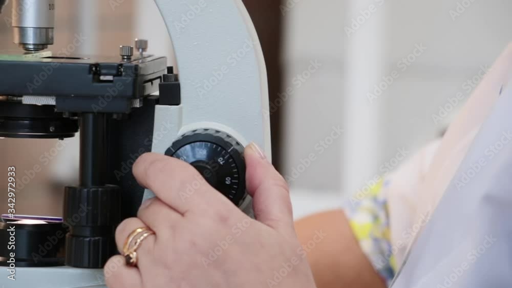 In close-up, the fingers slowly and gently rotate the microscope handle ...