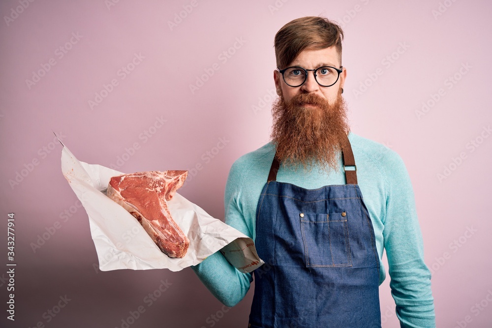 Redhead Irish butcher man with beard holding raw beef steak over pink ...