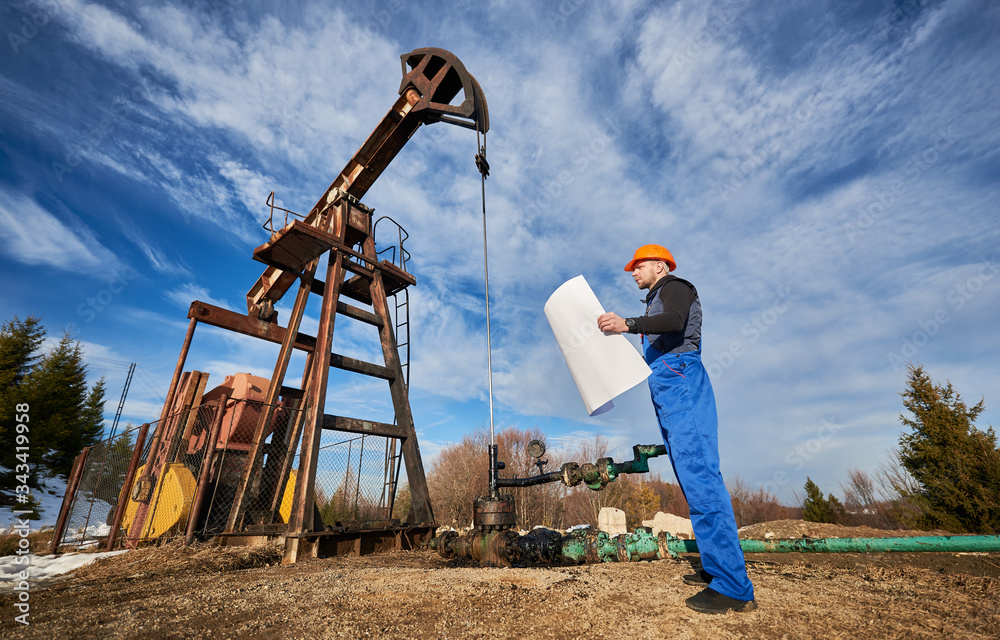 Petroleum worker holding plan of oil field and looking at oil pump jack ...