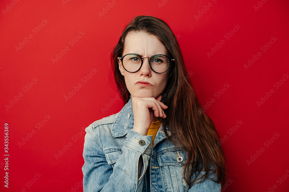 Young woman in glasses curves lips and frowns eyebrows, keeps hand ...