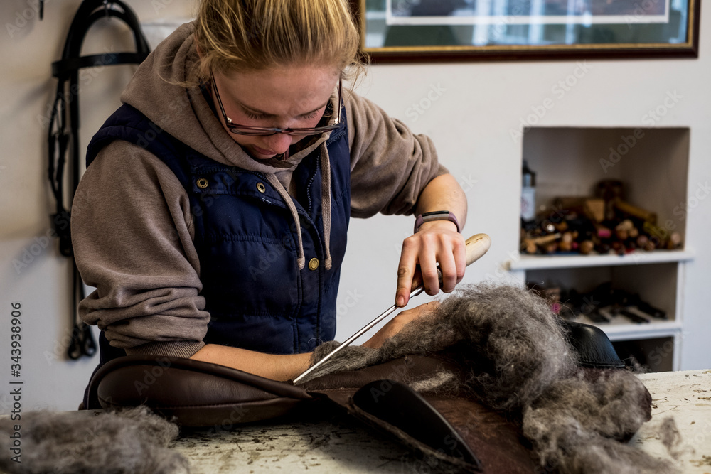 Female saddler standing in workshop, stuffing leather saddle with horse ...