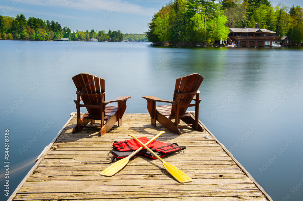 Two Adirondack chairs on a wooden dock facing the blue water of a lake ...