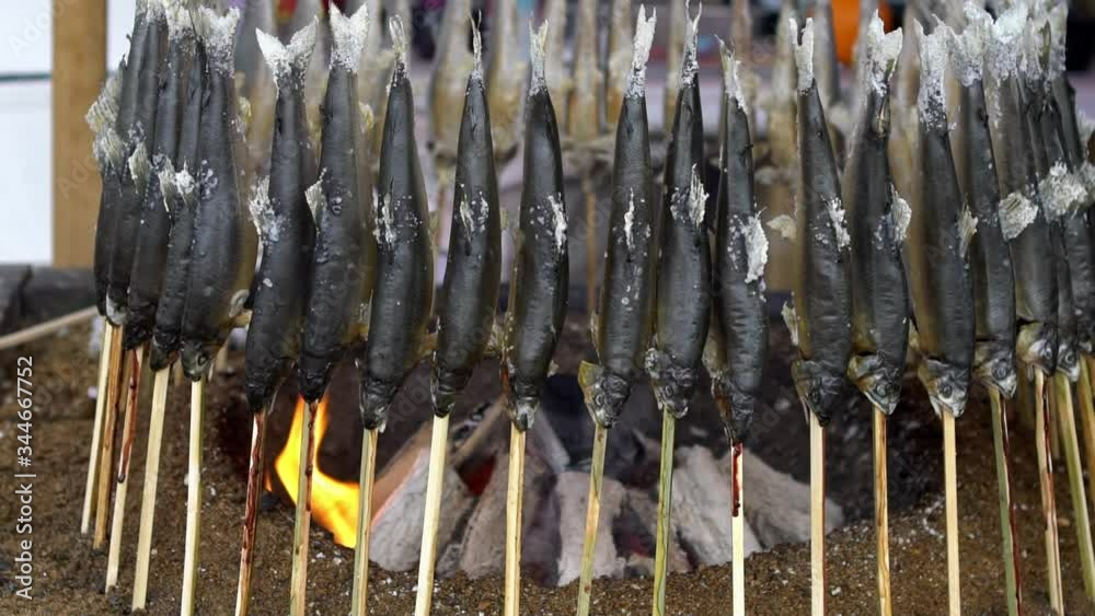 Slow motion of grilled fish next to coals on sticks. Vendor cooking a ...