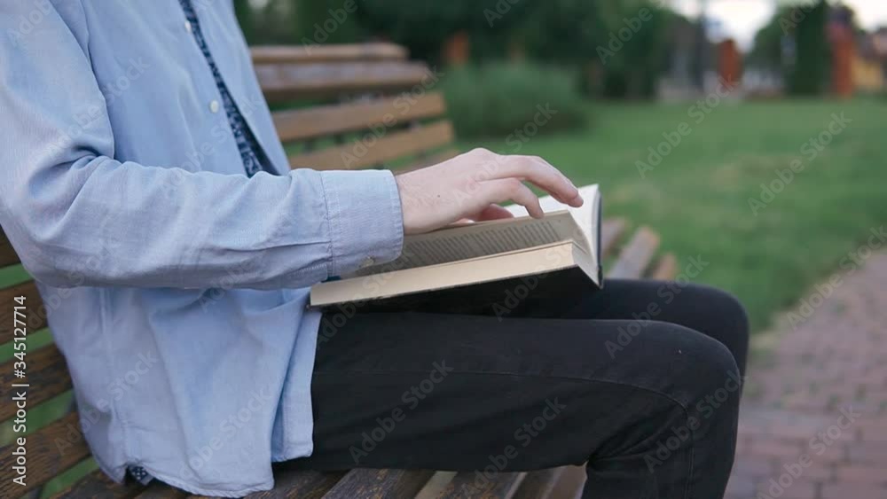 A man sitting on a bench in a park and reading a book, turning the page ...