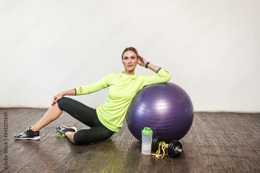 Slim beautiful woman in tight sportswear sitting on floor with big rubber gym ball, tonic drink ...
