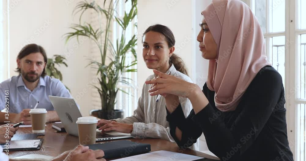 Smiling young arab female employee in hijab sitting at table with ...