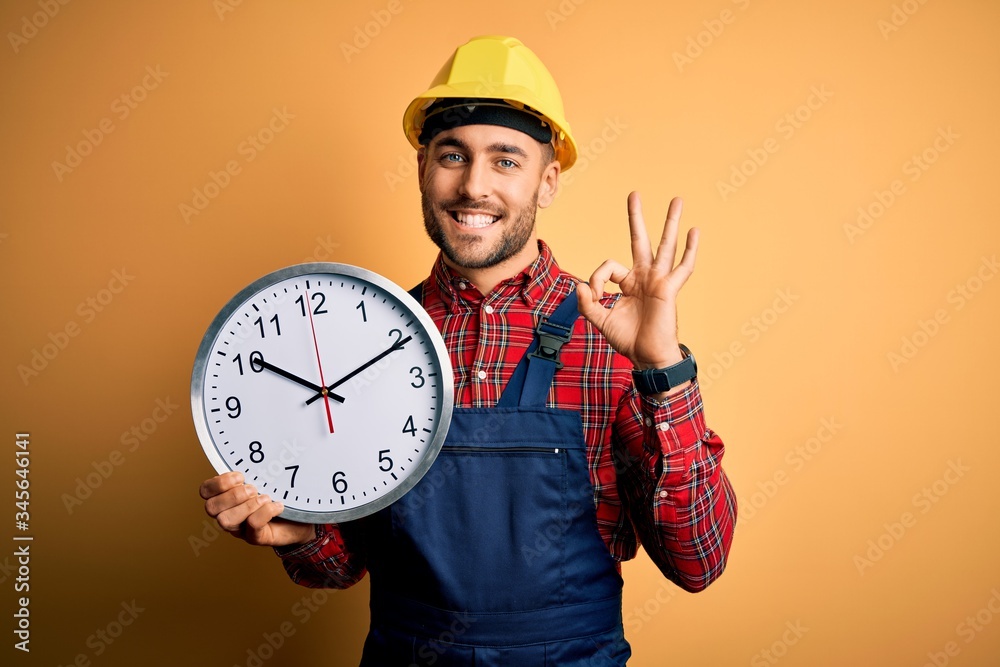 Young builder man wearing safety helmet holding big clock over yellow ...
