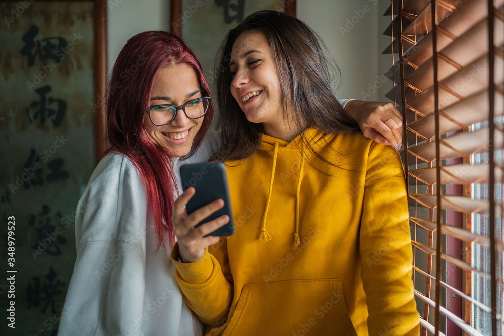 Two attractive young girls next to a window consulting the mobile phone ...