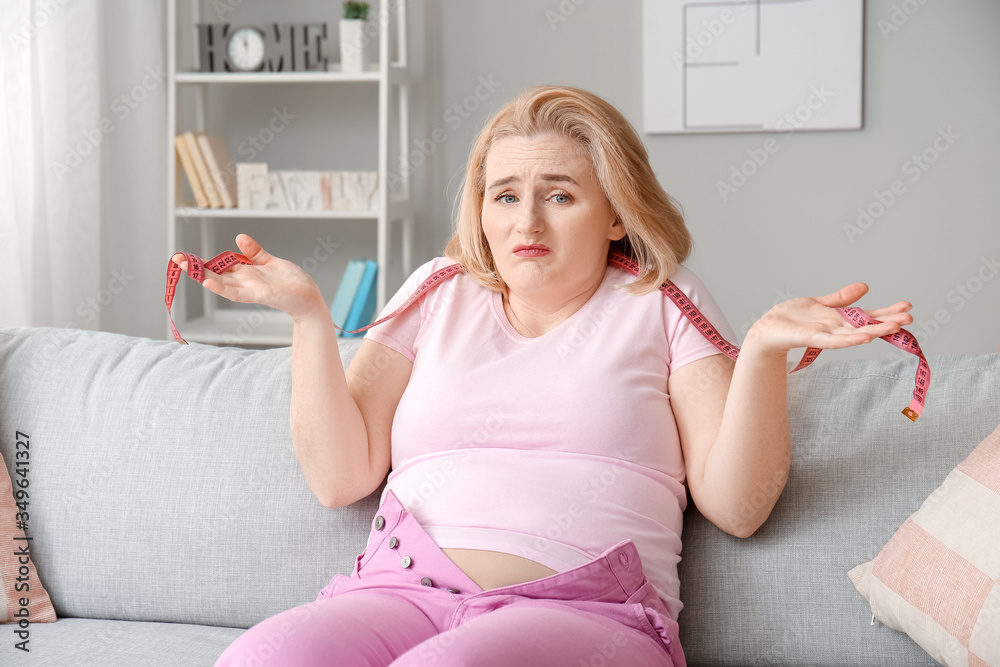 Sad overweight woman in tight clothes and with measuring tape at home ...