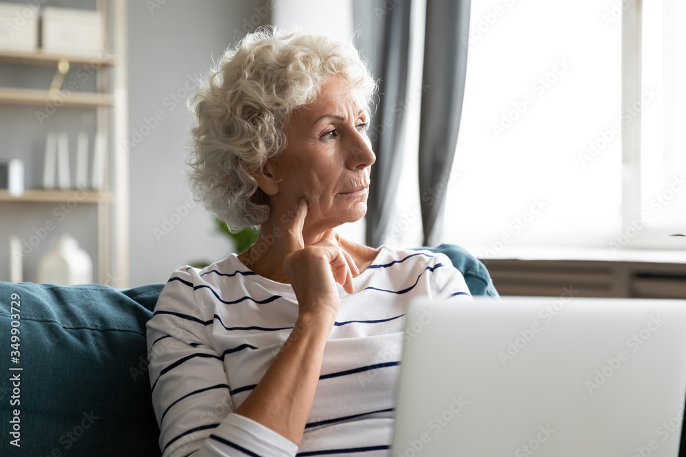 Thoughtful mature senior woman sitting on couch with laptop on lap ...