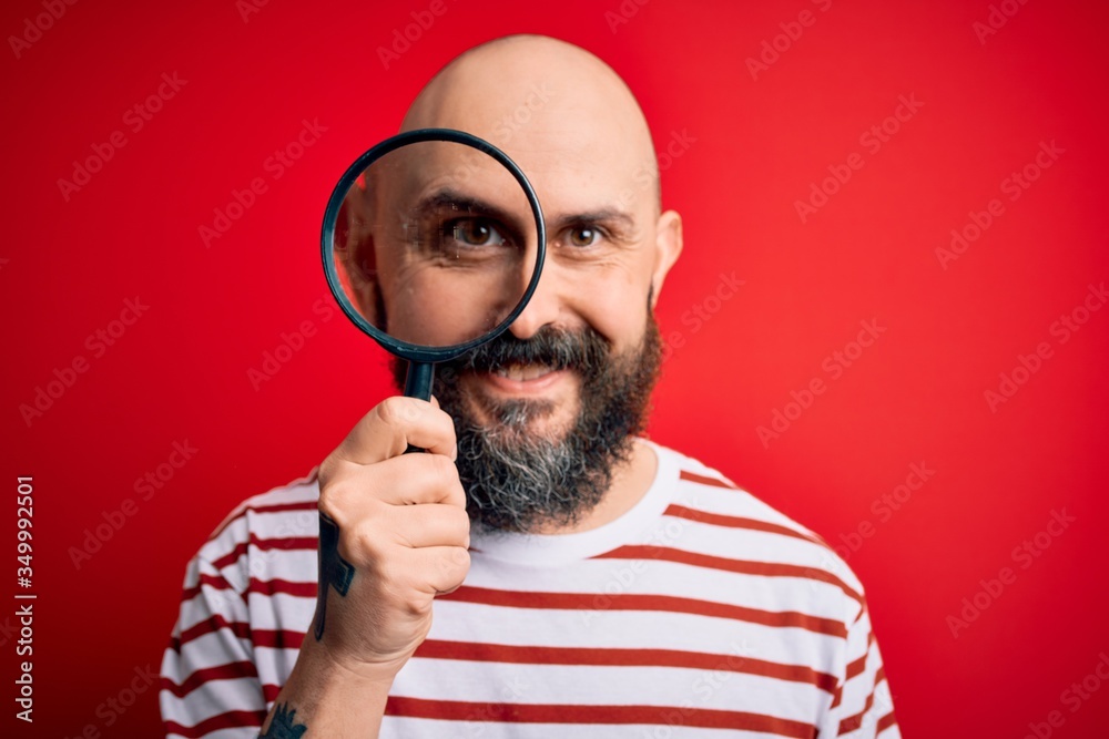 Handsome detective bald man with beard using magnifying glass over red ...