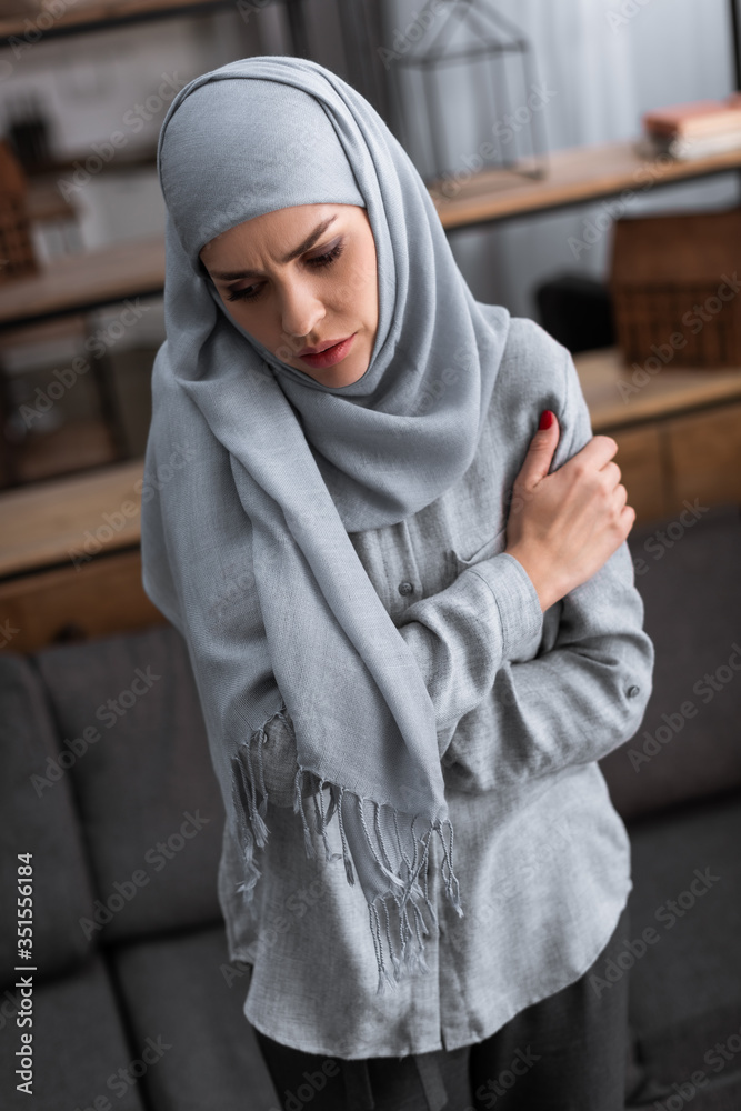 sad muslim woman in hijab touching arm in living room, domestic ...